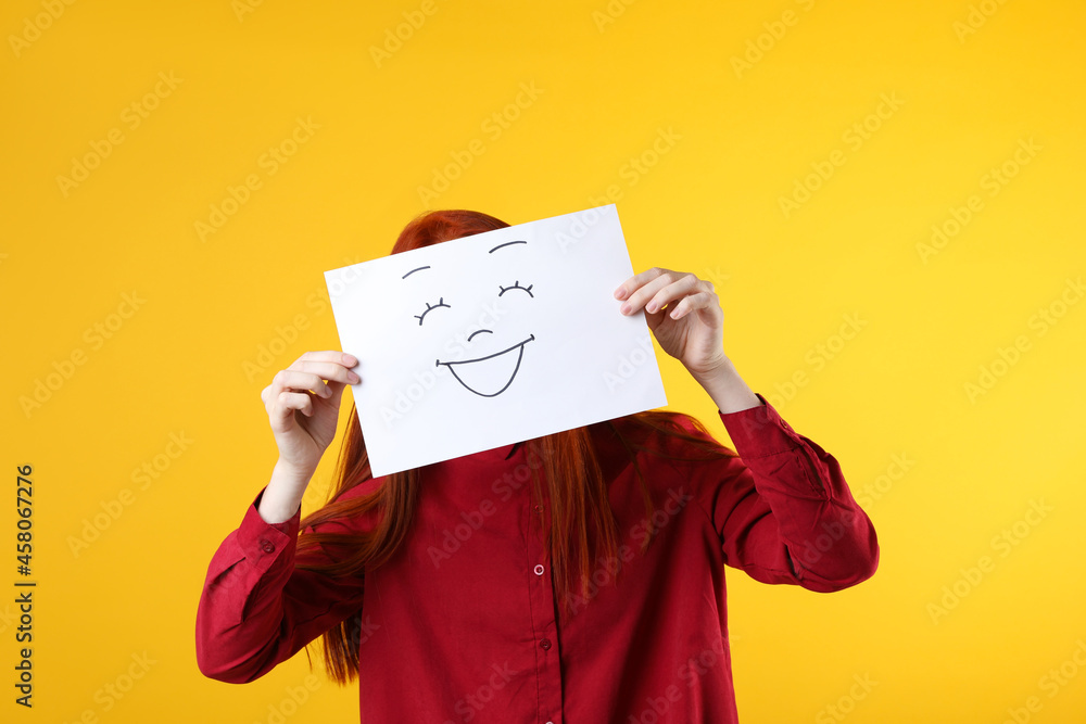 Woman hiding behind sheet of paper with happy face on yellow background ...