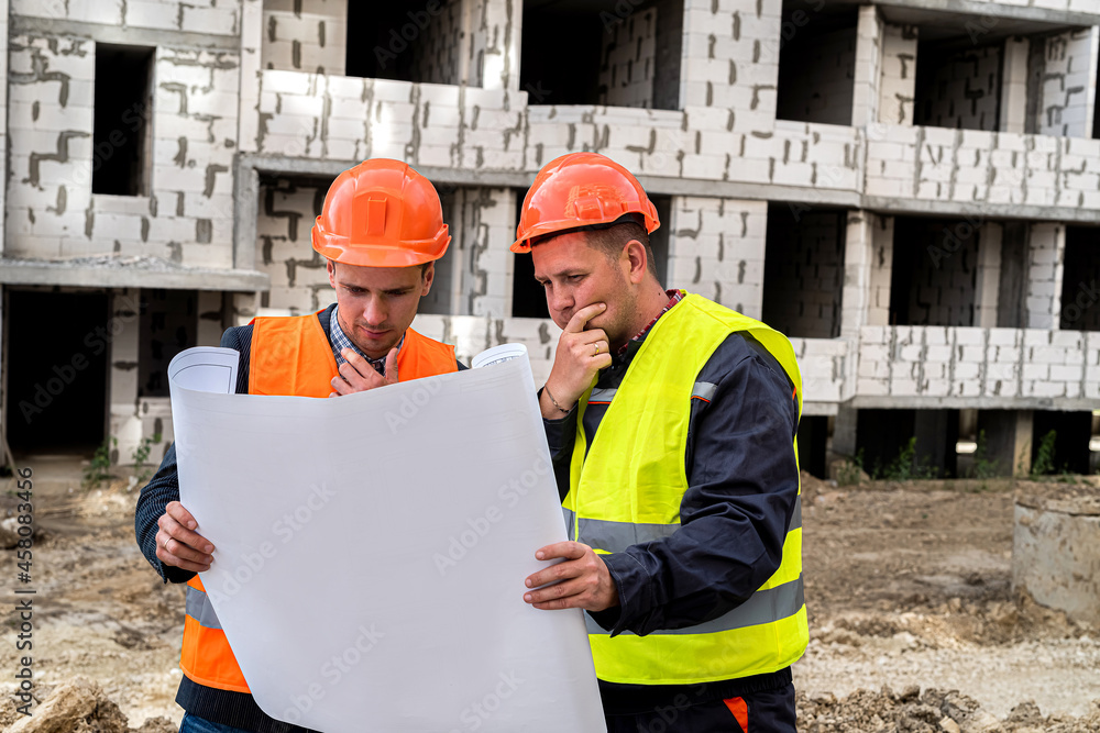 two young workers in uniform on construction site stand with plan and ...