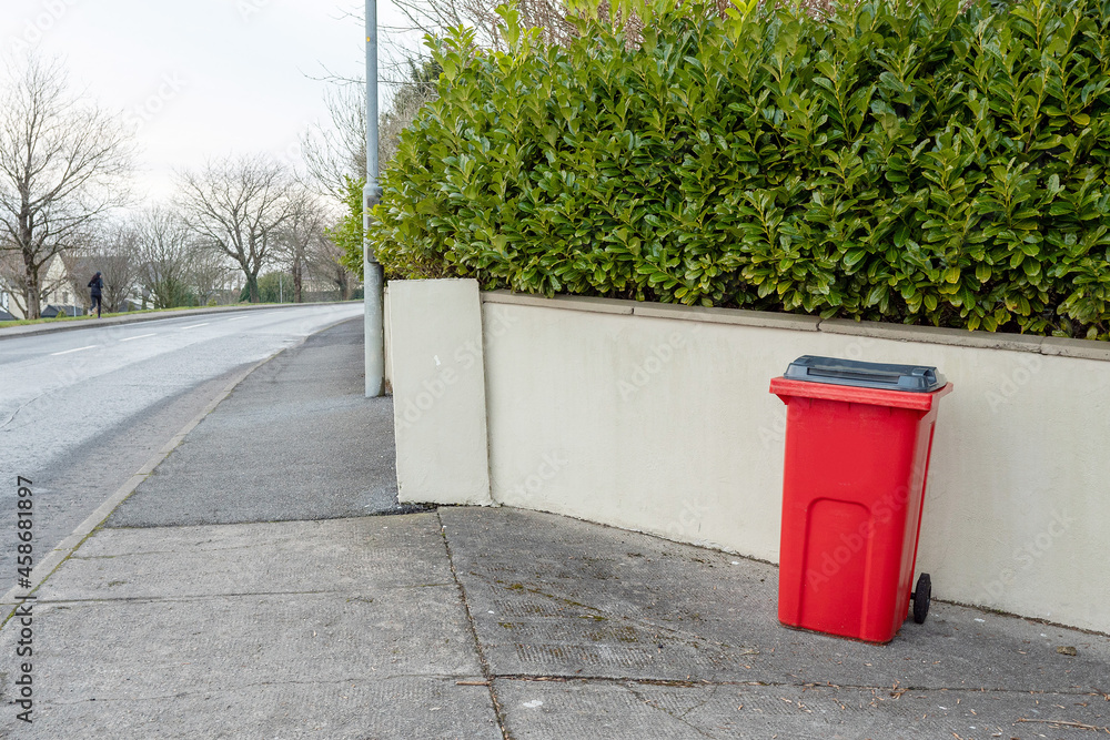 Red plastic wheelie bin out in a street ready for collection by a white ...