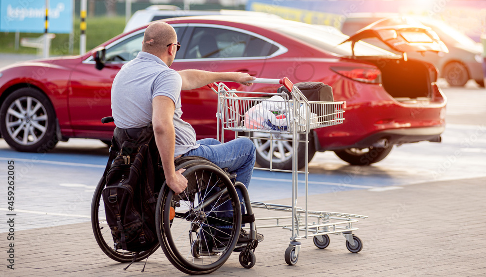Adult disabled man in a wheelchair pushes a cart towards a car in a ...