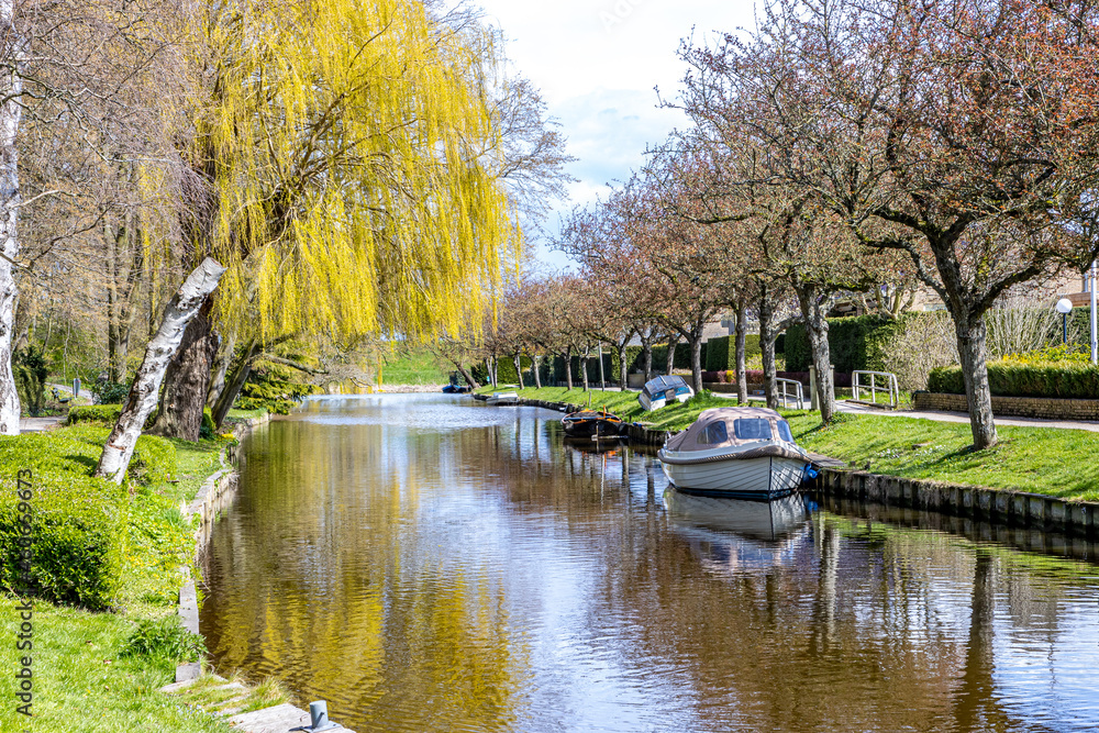 Canal surrounded by grass, bare trees and weeping trees, boats anchored ...