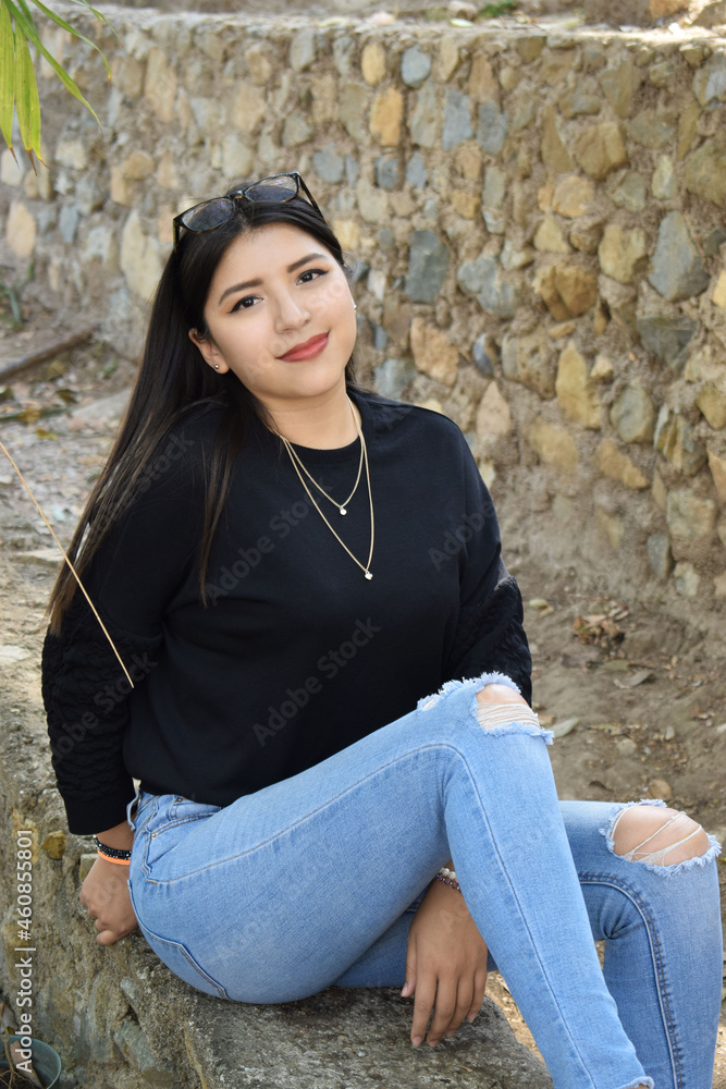 Mujer joven latina, sentada sobre una banca de piedra, usando una blusa negra y pantalón azul ...