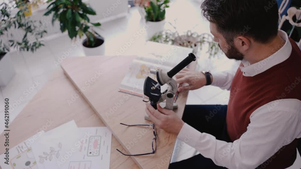 Male teacher using microscope and looking at biology textbook in modern ...