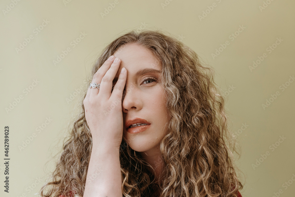 Young Woman With Curly Hair Covering Eye With Hand Against Wall Stock