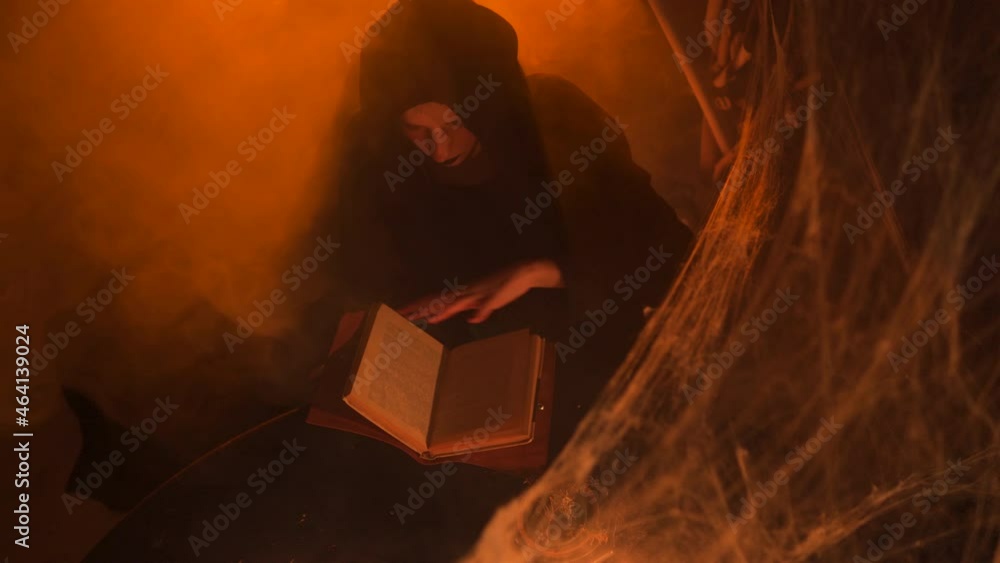 A young witch in hood does black magic ritual using the Witch's book ...