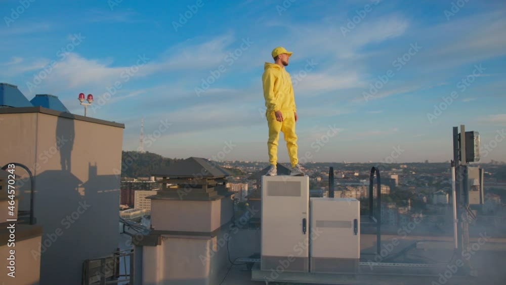 Young hip hop dancer stylish guy in yellow streetwear walking on rooftop building and dancing ...