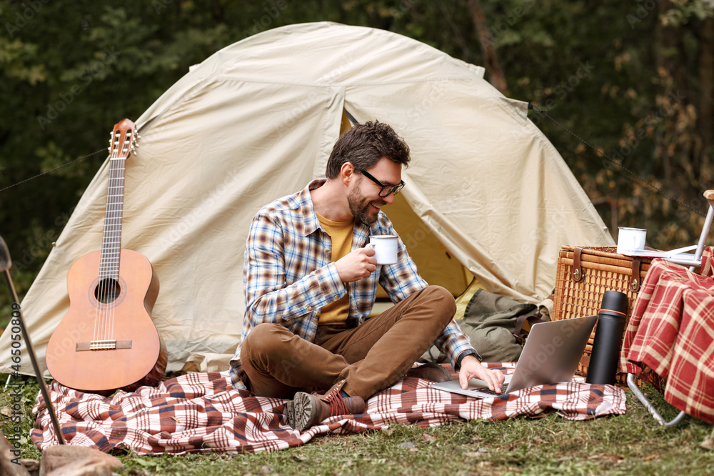 Smiling man sitting near tent with laptop computer and working remotely ...