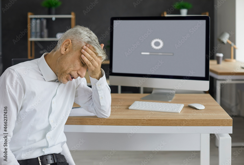 Unhappy, sad senior man sitting at office desk with computer with gear ...