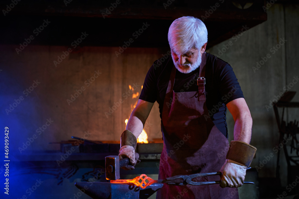 Caucasian man with grey hair and beard processing molten metal at forge ...