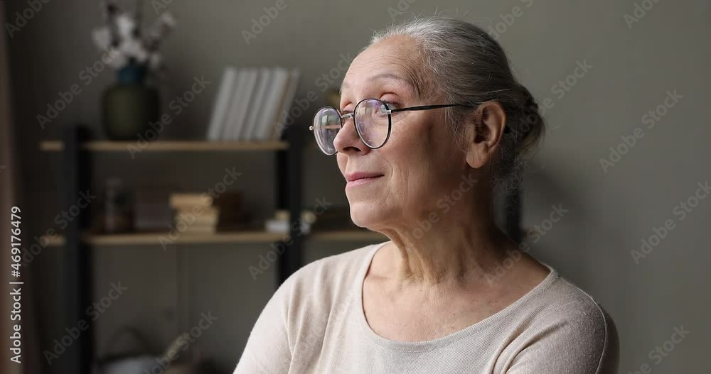 Thoughtful dreamy grey haired old age lady wear glasses stand at living ...
