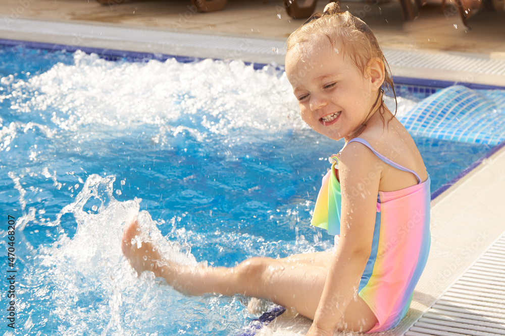 Little girl in rainbow swimsuit sit on the pool edge, swinging her feet ...