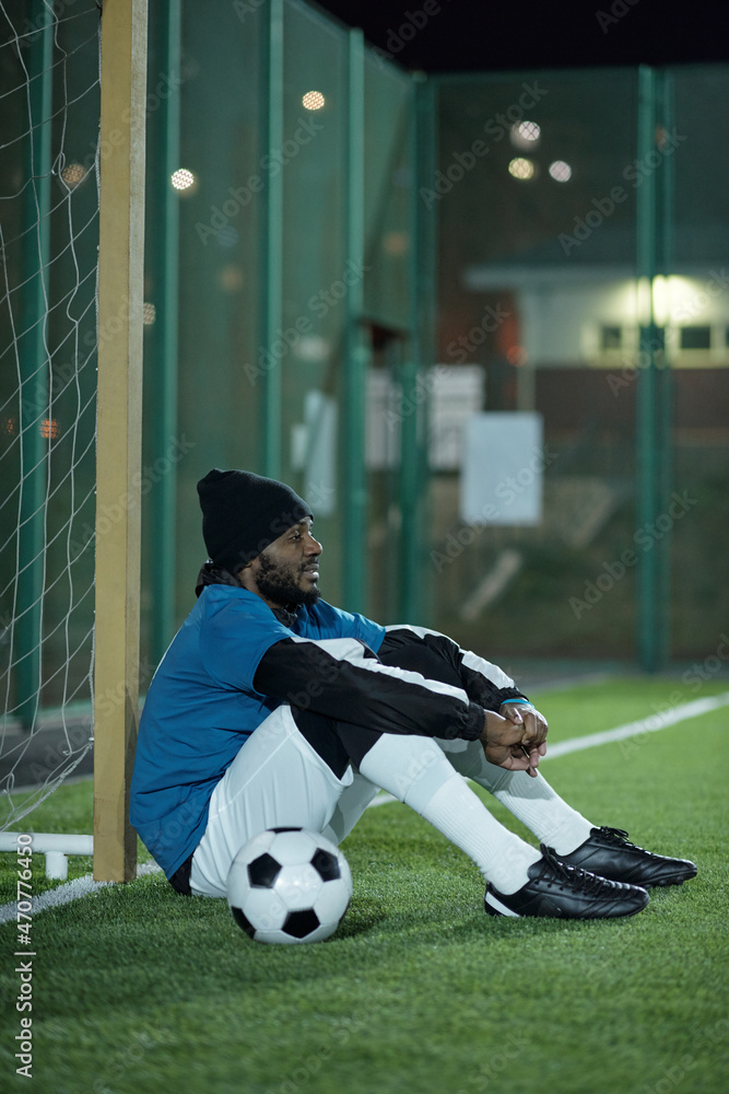 Young restful football player of African ethnicity sitting by net with ...