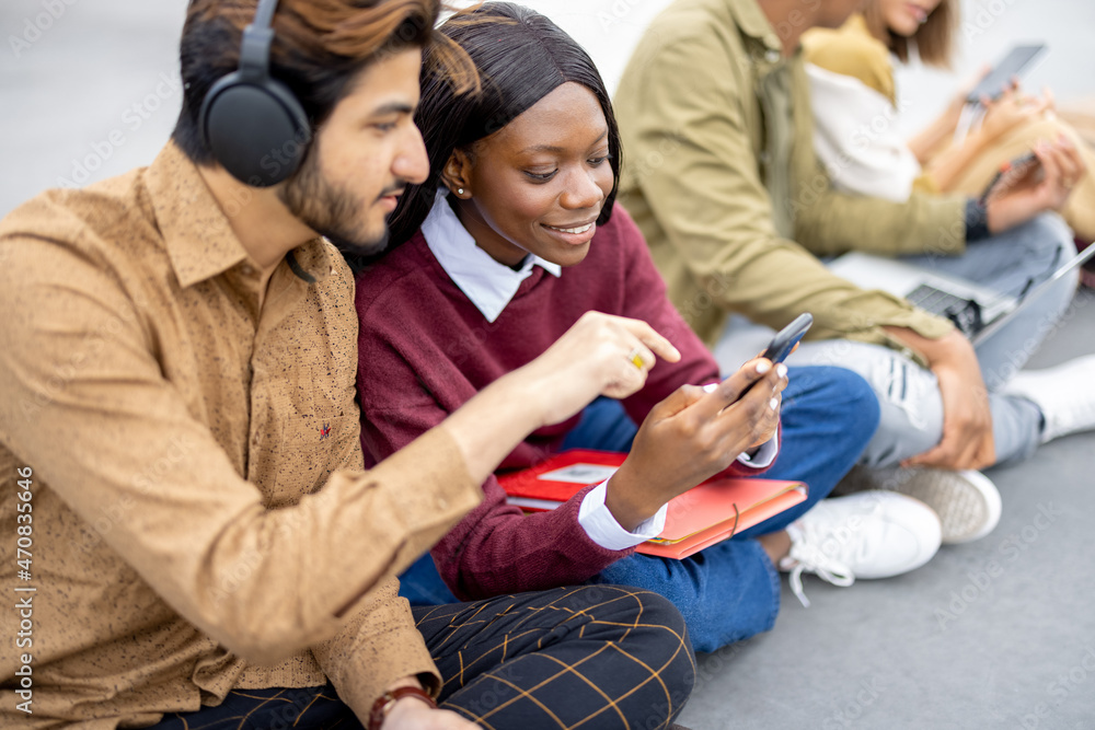 Multiracial students sitting and using digital devices on asphalt at ...