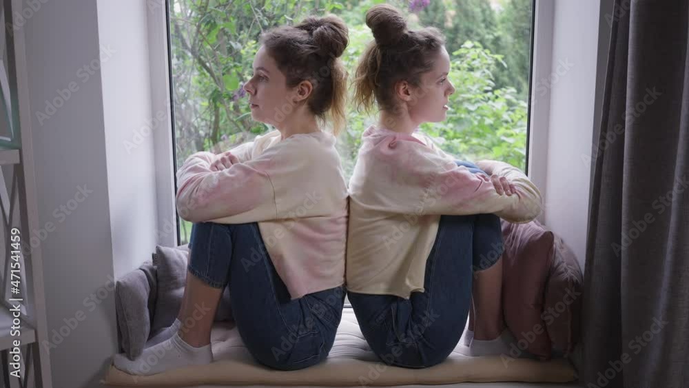 Side view two identical young women sitting on windowsill back to back ...