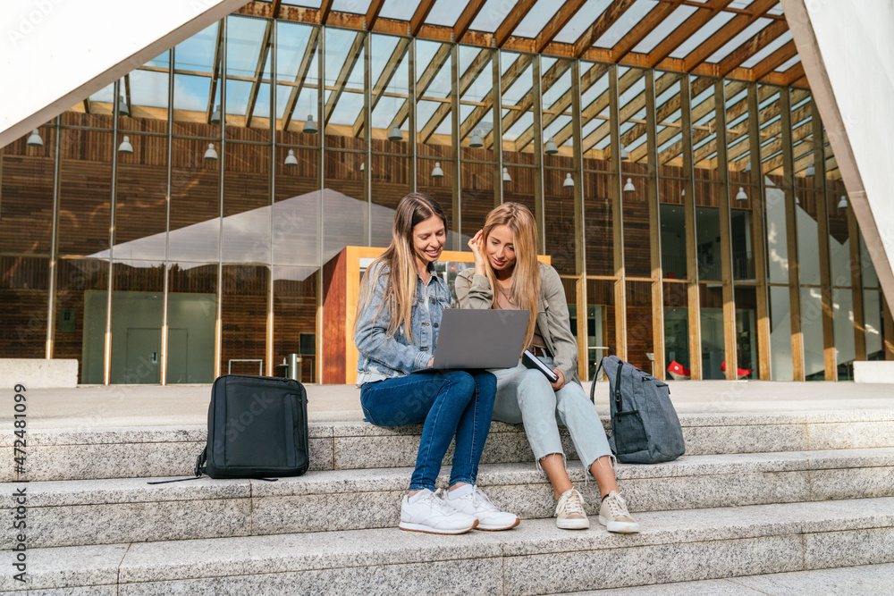 Two college classmates using a laptop sitting on the staircase of the ...