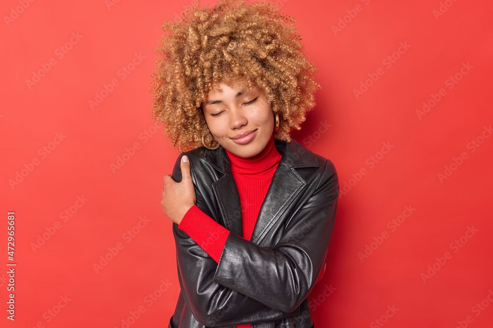 Indoor Shot Of Curly Haired Woman Embraces Herself Keeps Eyes Closed