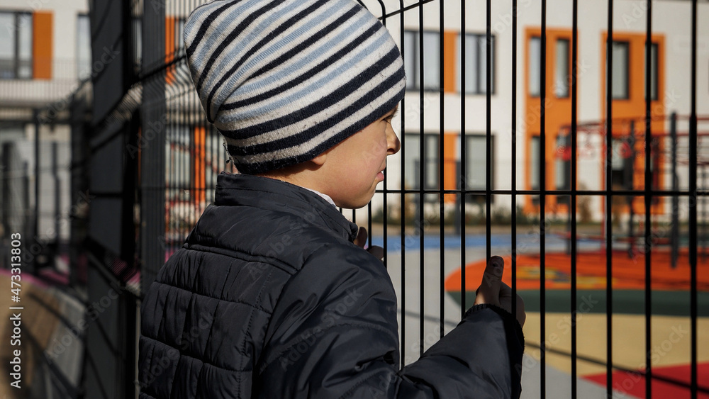 Little upset boy looking through metal fence on modern school and ...