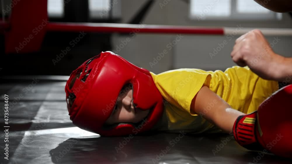 Little boy in boxing gloves and soft helmet falls down on the ring in ...