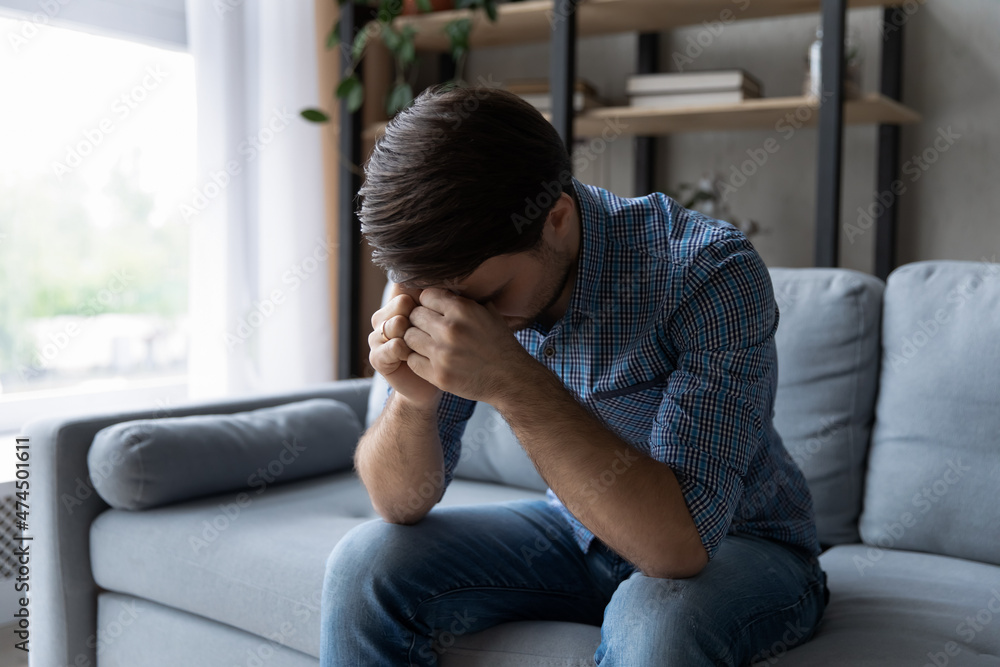 Sad depressed millennial man sitting on sofa in living room with head ...