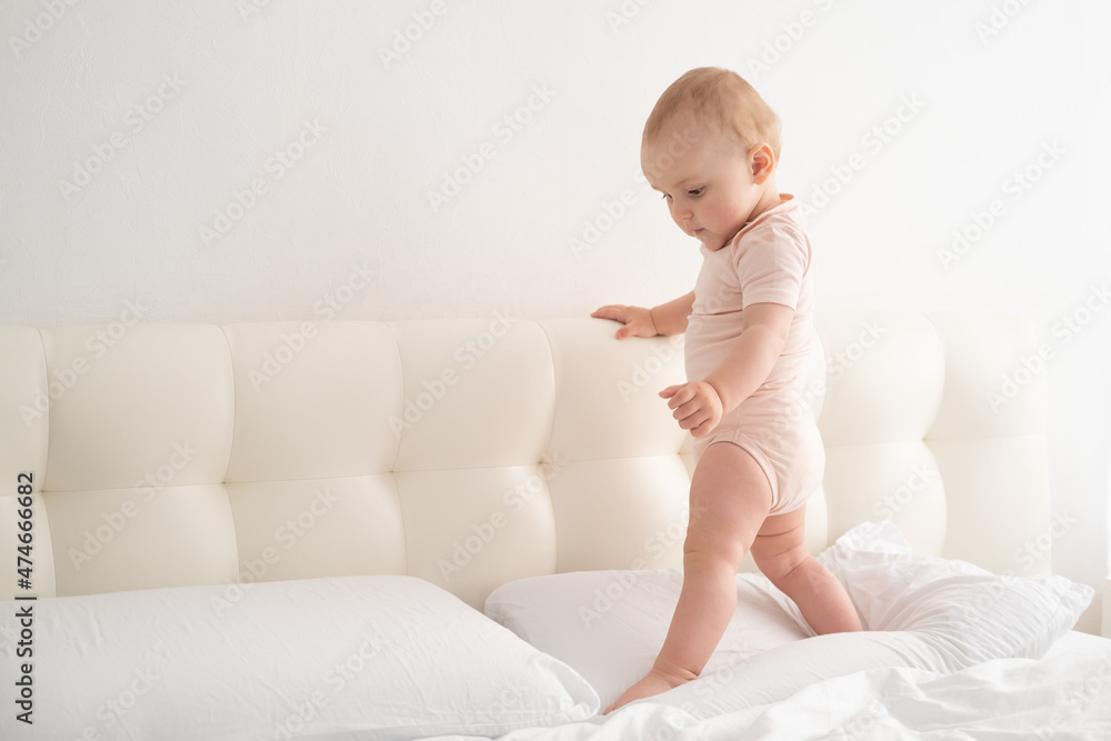 Cute baby girl in bodysuit stand holding bed frame, learning to walk on ...