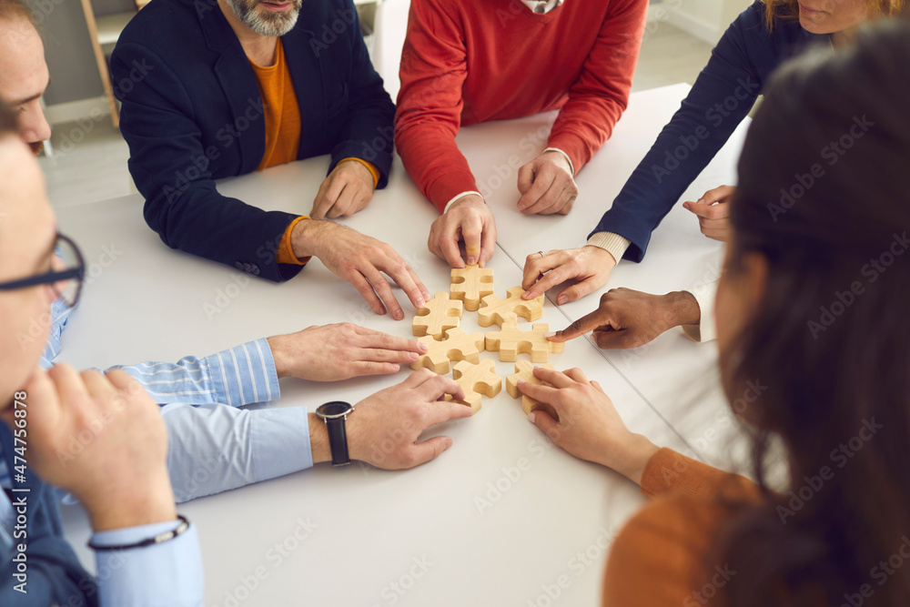 Team of business people sitting around office table and joining wooden jigsaw puzzle pieces ...