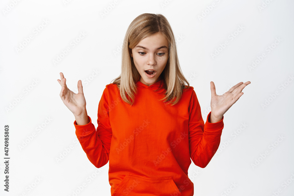 Surprised and shocked little blond girl looks down startled, seeing ...