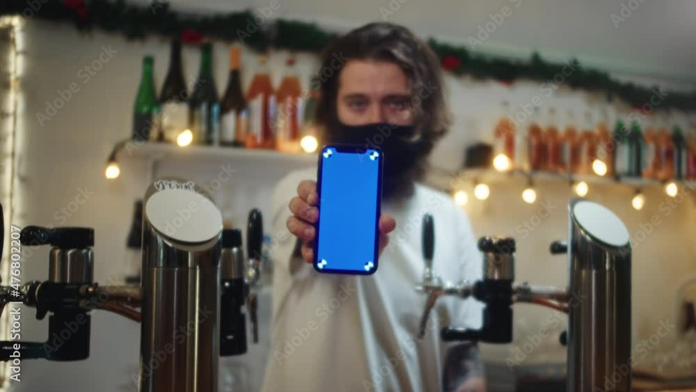 Barman in mask showing blue screen on phone. Bearded man behind bar ...