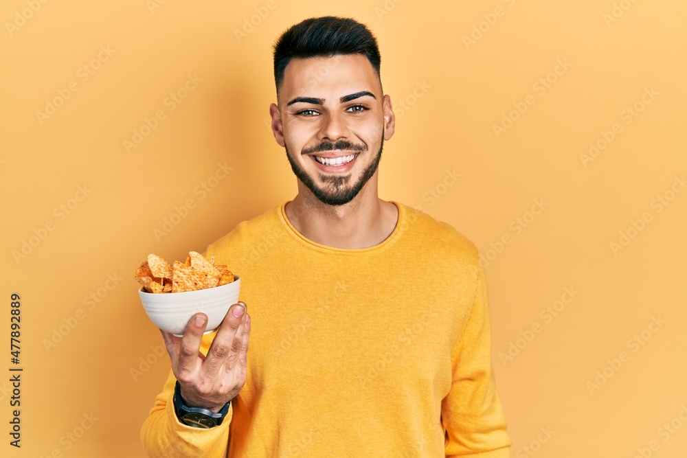 Young hispanic man with beard holding nachos potato chips looking ...