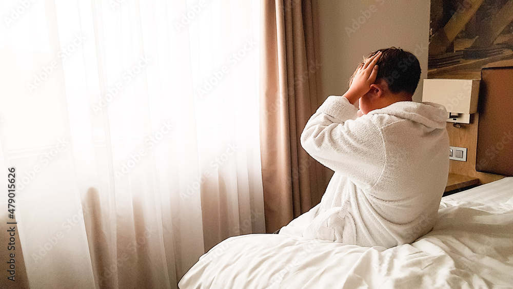 the teenager holds his head and sits on the bed confused. headache ...