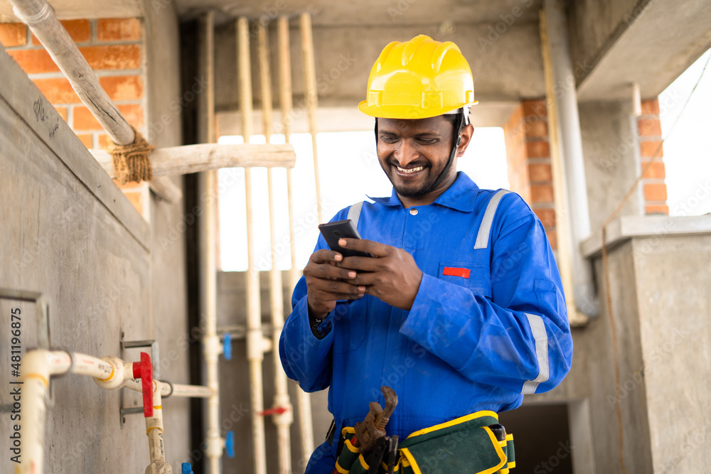 smilling young indian plumber or repairman busy by using mobile phone ...