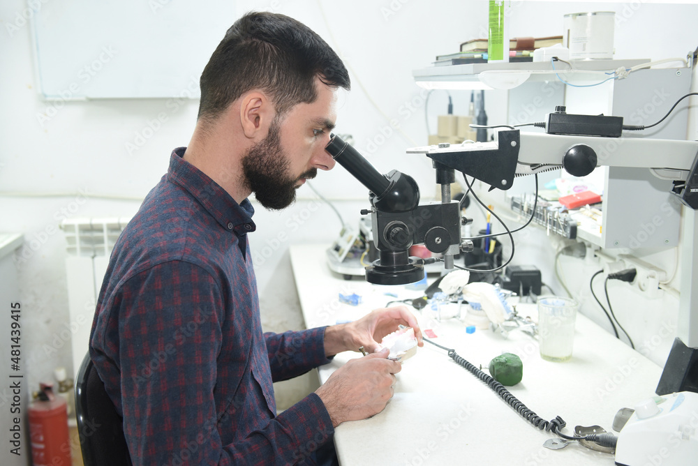 A young man looks through a microscope examining repairs dentures or a ...