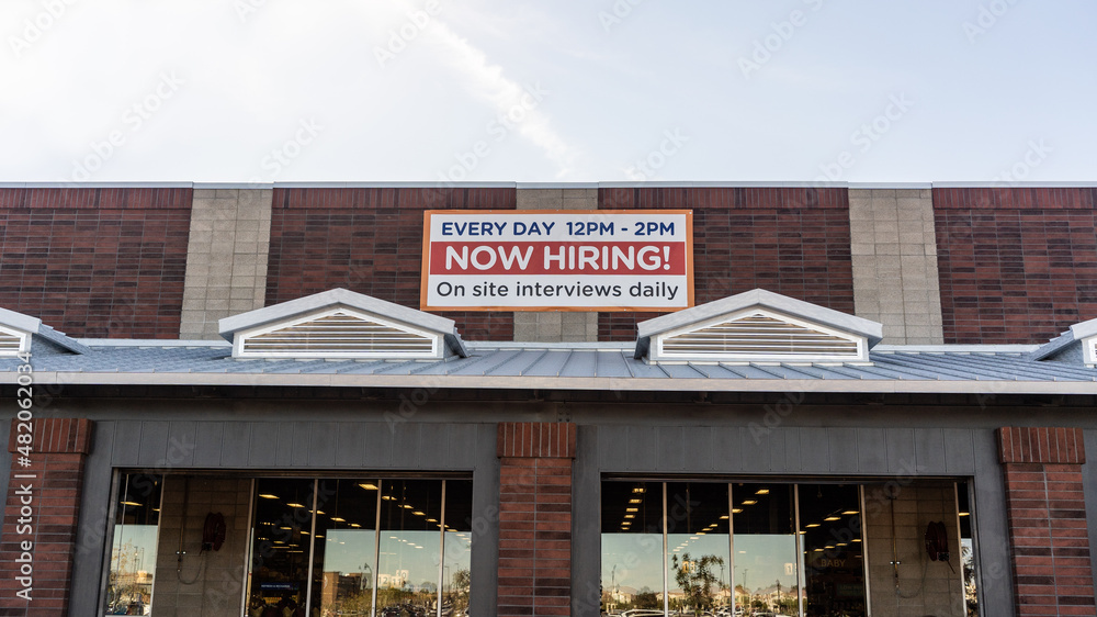 Now Hiring sign on outside of red brick retail store with blue sky ...