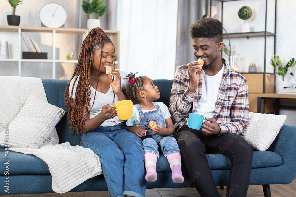 Young parents and their little daughter resting on sofa with cups of ...