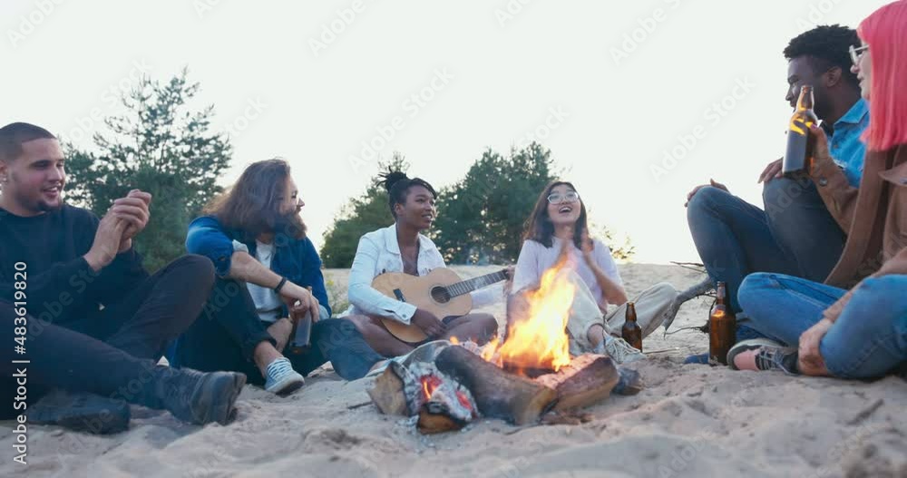 Friends are having fun together on beach sitting on sand by campfire ...