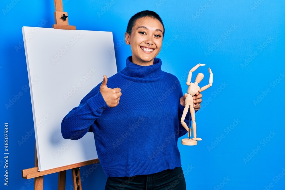 Beautiful hispanic woman with short hair holding manikin close to easel ...
