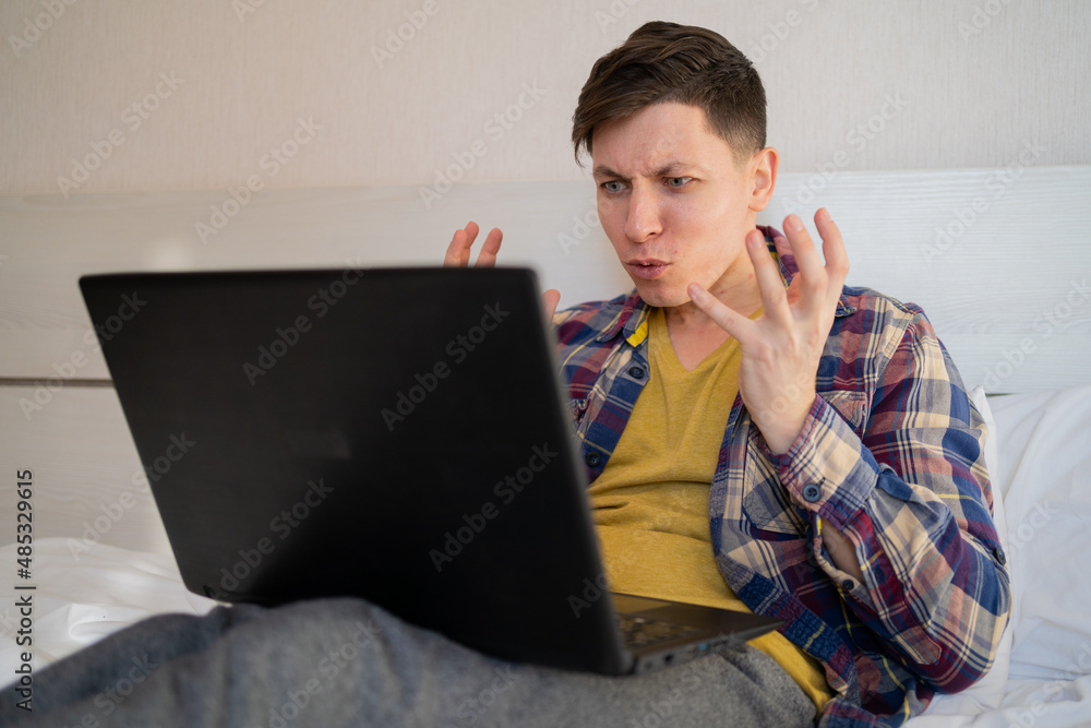 Irritated man sitting on bed looking at laptop screen, arms ...