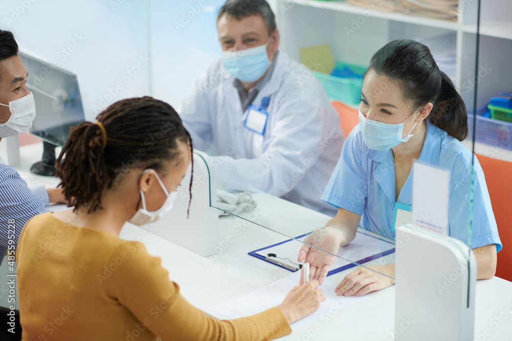 Medical nurse behind glass screen helping patient in protective mask to ...