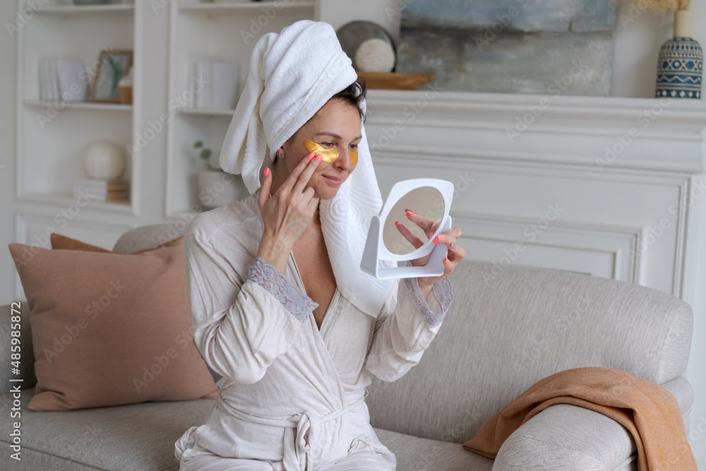 Woman in with towel on her head looks in mirror and applies golden ...