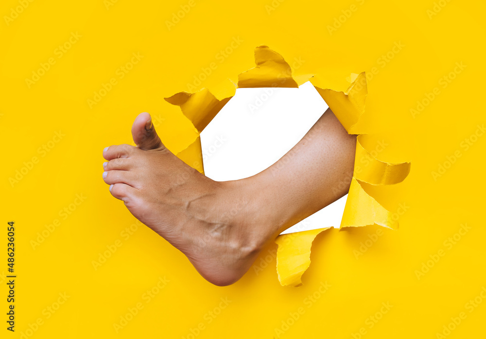 A left woman's foot appears in a torn hole in yellow paper and tries to ...