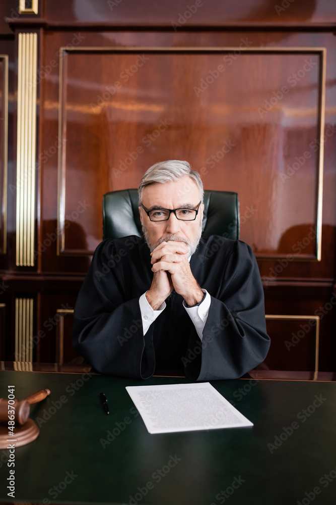 grey-haired judge in robe and eyeglasses sitting with clenched hands ...