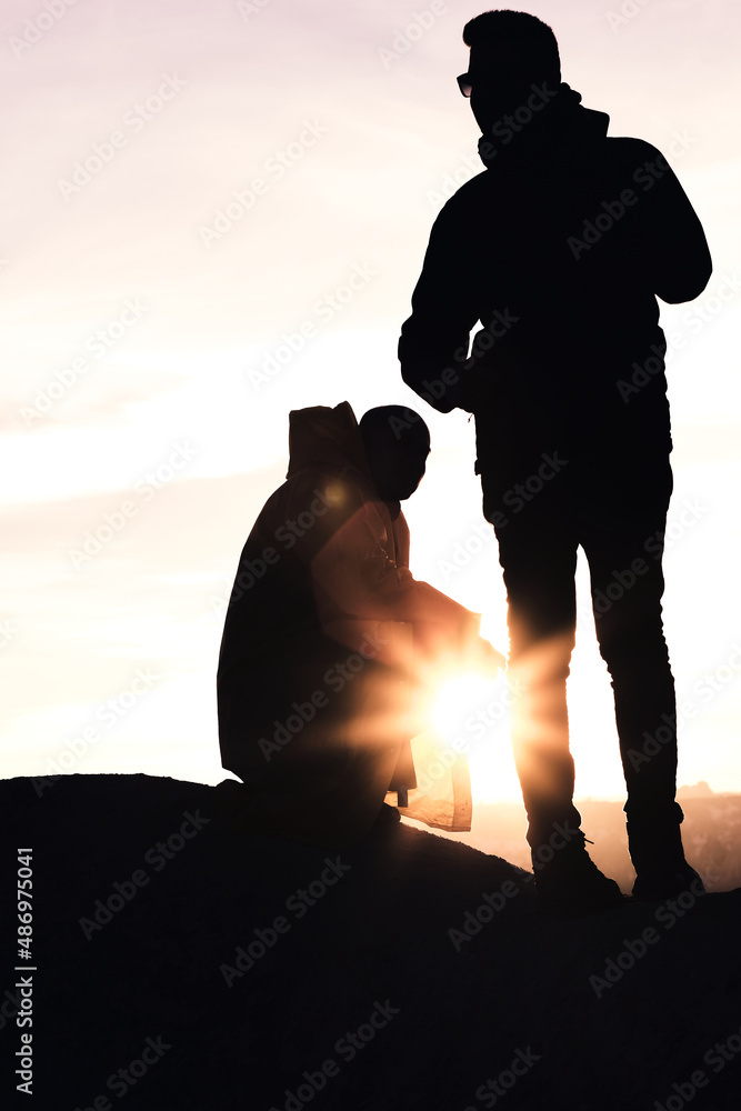 Silhouettes of two men, one crouching and the other standing, on a hill ...