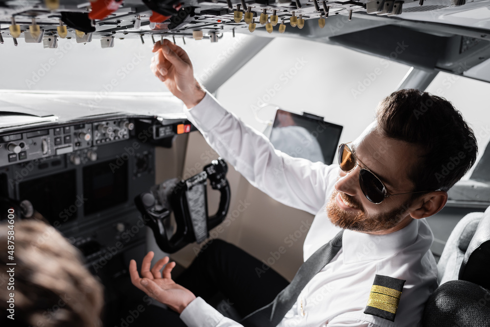 cheerful pilot in sunglasses reaching overhead panel near co-pilot in ...
