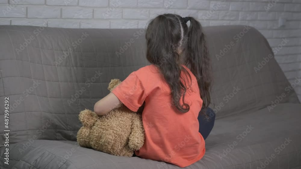 Angry child with toy. An angry little girl hugs her teddy bear and sits ...