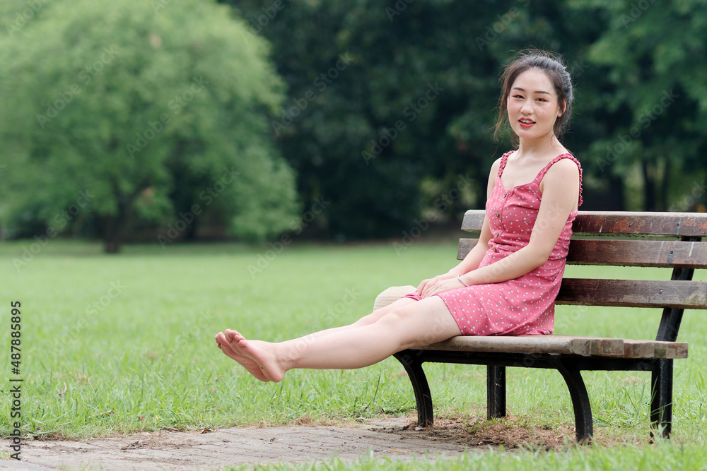 Portrait Of Young Asian Woman Sitting On Bench On Grass Field With Barefoot In Forest Park