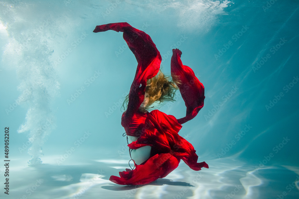 Attractive red-haired young woman swims beautifully underwater in a red ...