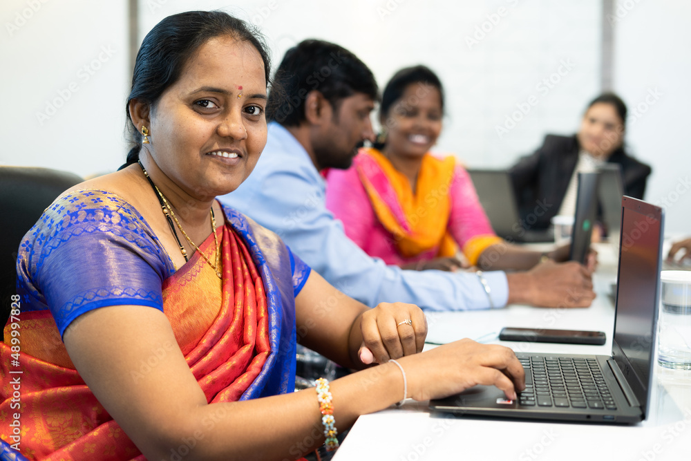 smiling Indian woman looking at camera while busy working on laptop at ...