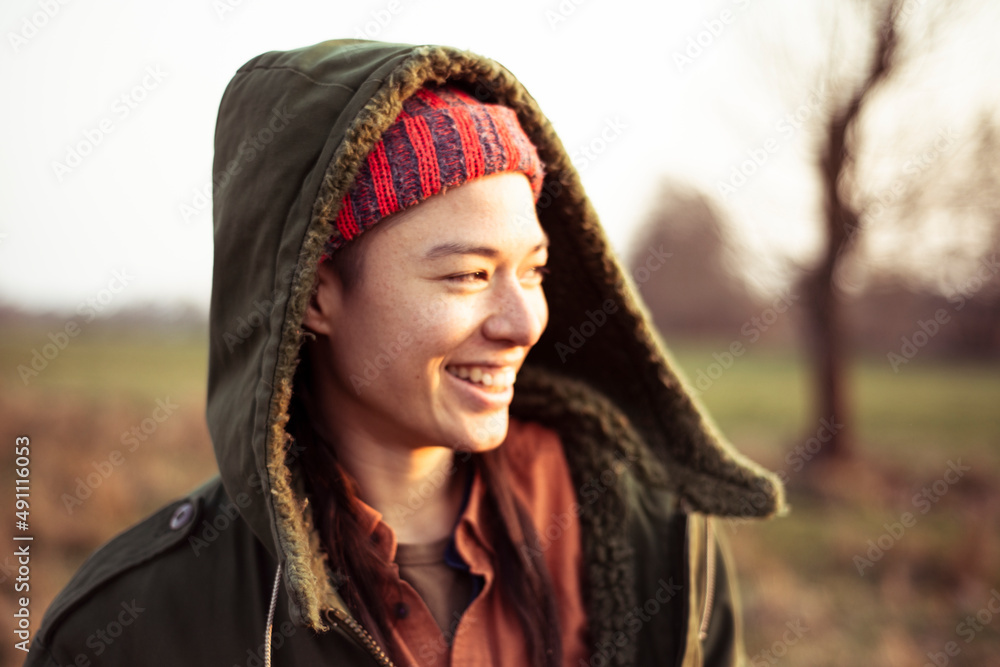 non-binary person in warm jacket hood smiles in afternoon sun outdoors ...
