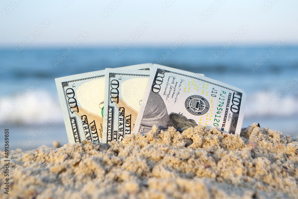 Three one hundred dollar bills half buried in sand on sandy seashore ...