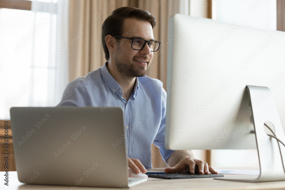Smiling young man software developer sit at table work on two computer ...