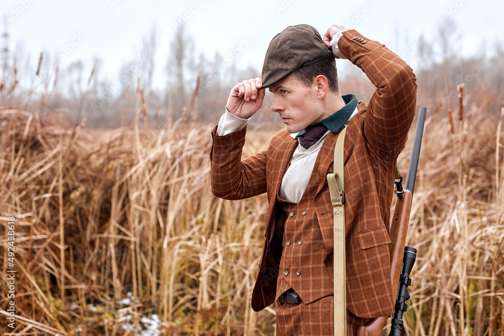 Hunter man adjust the hat, in trendy suit. Hunter with hunting gun in ...